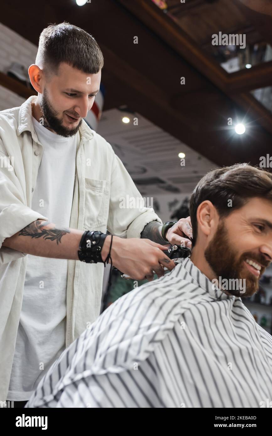 Smiling tattooed barber trimming neck of bearded client in barbershop ...