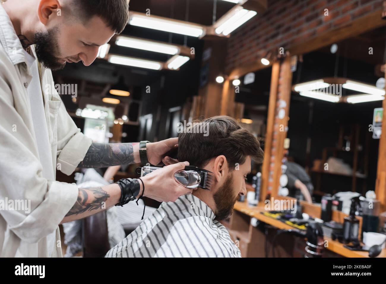 Side view of tattooed barber holding trimmer near client in barbershop ...