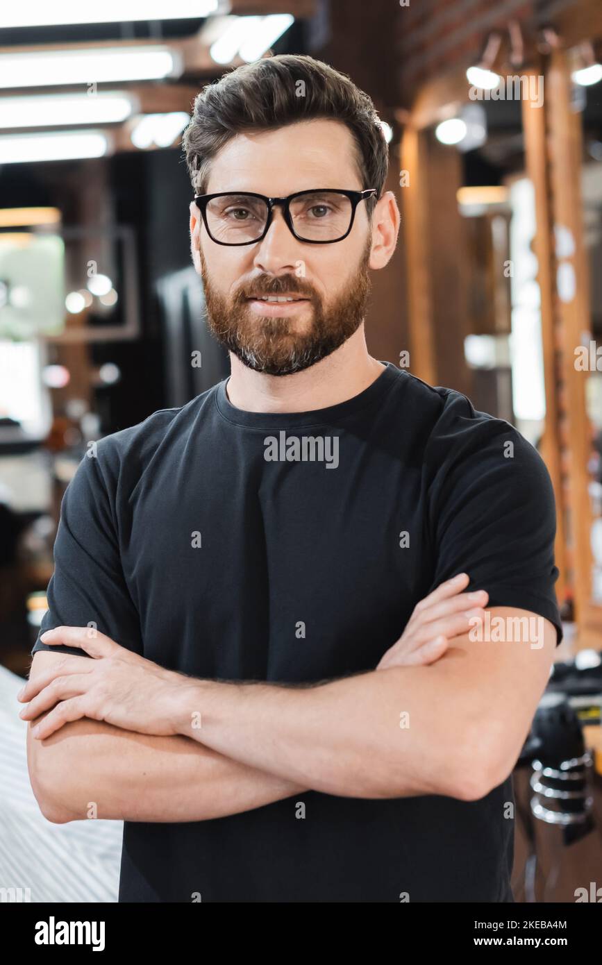 Bearded barber in eyeglasses crossing arms and looking at camera in ...