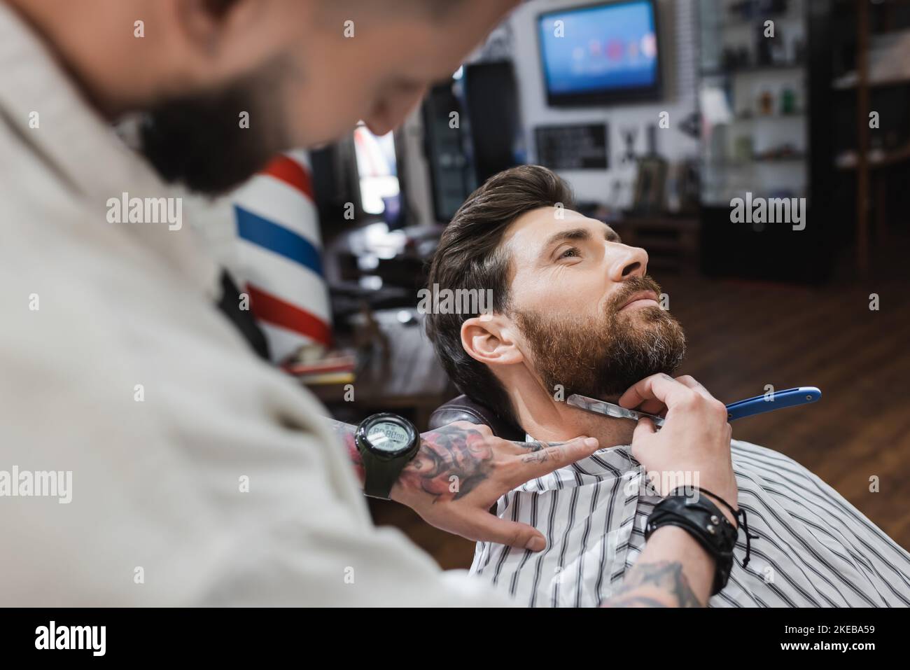 Tattooed barber shaving bearded of man with straight razor in ...