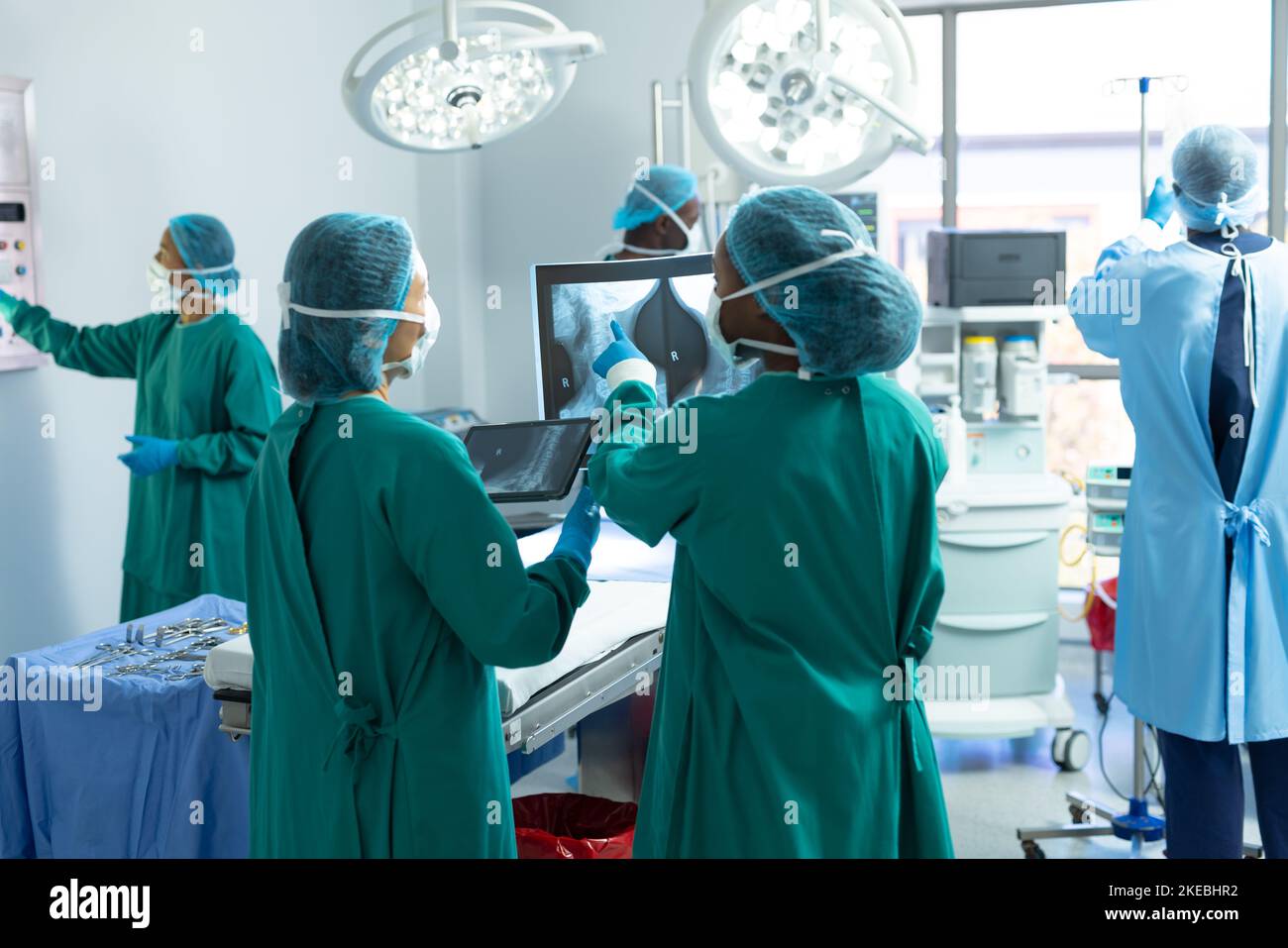 Rear view of two diverse female surgeons discussing x-ray in operating theatre, with colleagues ...