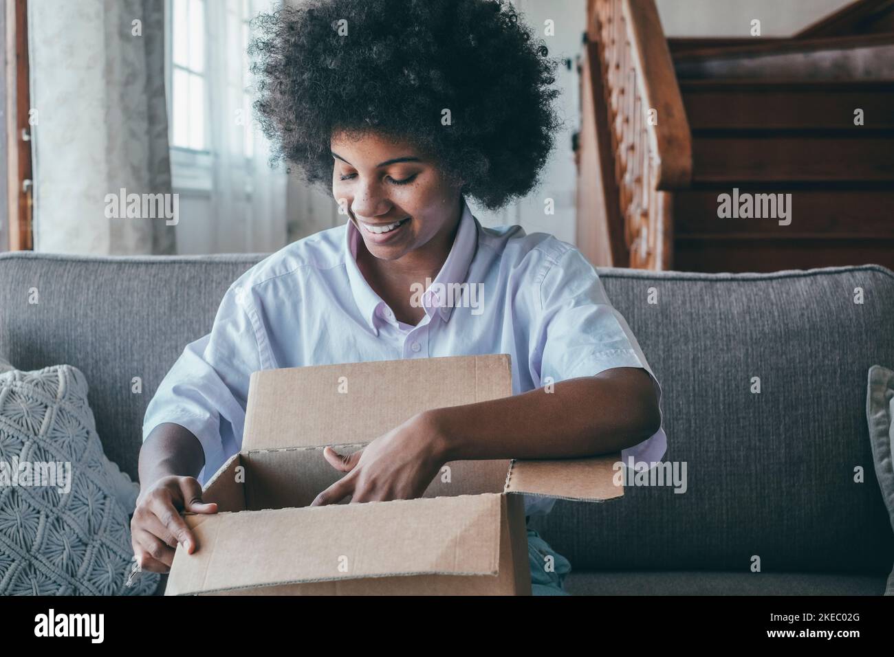 African woman sitting on sofa in living room hold on lap big carton box unpacking parcel ...