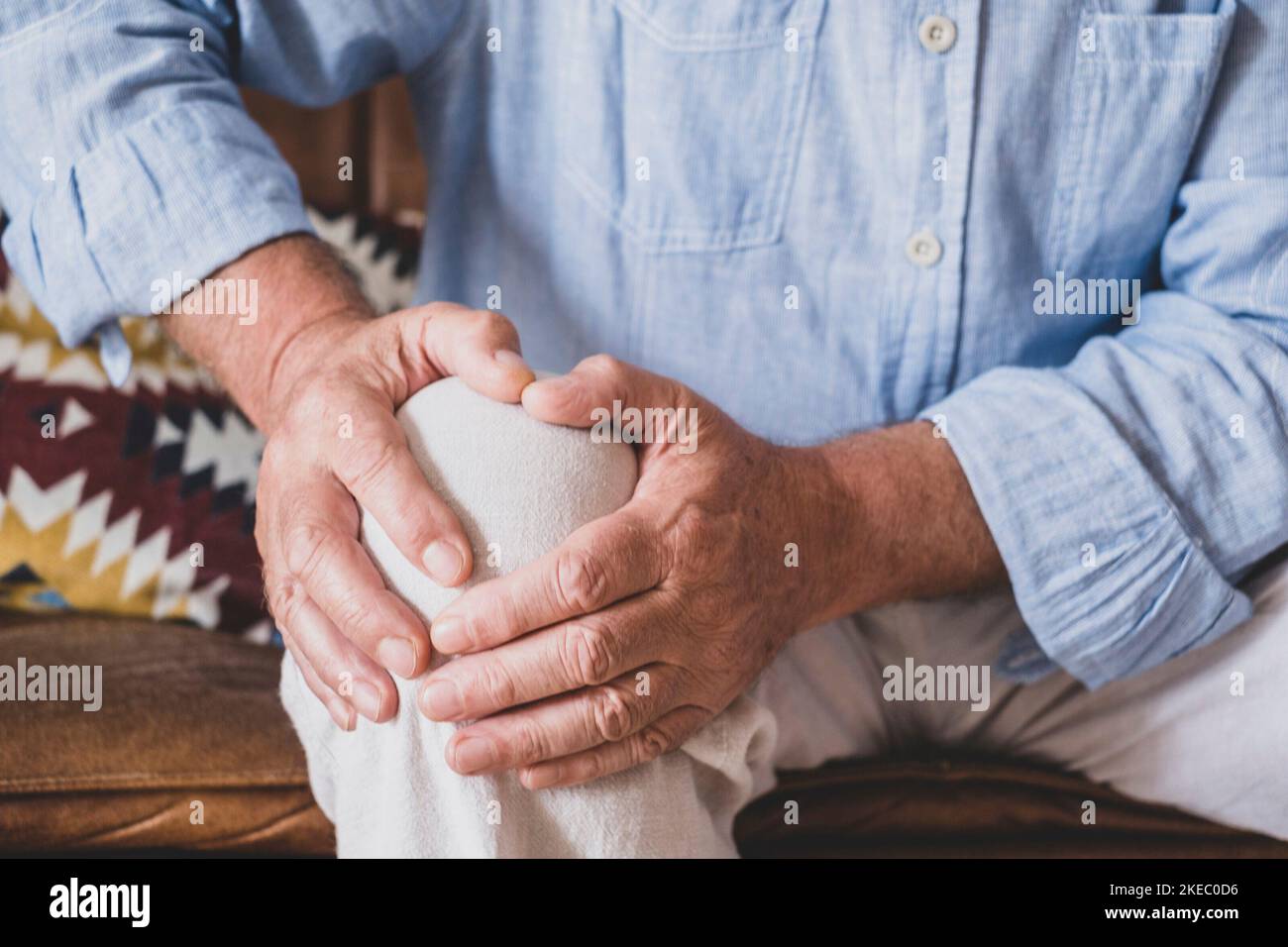 Close Up Of Senior Old Man Facing Knee Problem Sitting On Sofa Holding Knee At Home Elderly