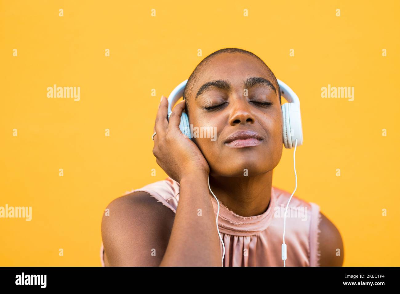 Dark skinned woman listening to music on headphones with eyes closed over yellow background ...