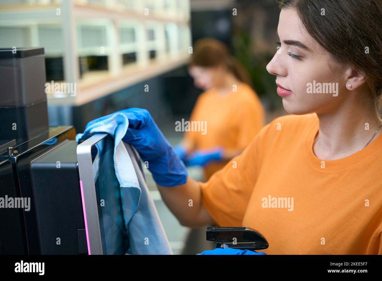 Woman cleaner washes coffee machine, her colleague is in background ...