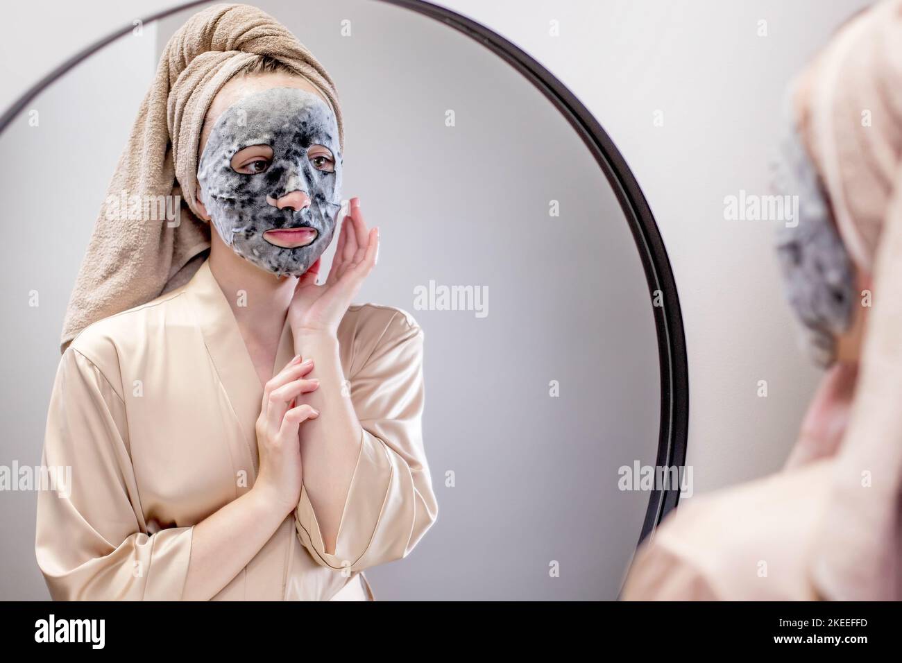 Young happy woman in towel making face mask and looking in mirror in ...