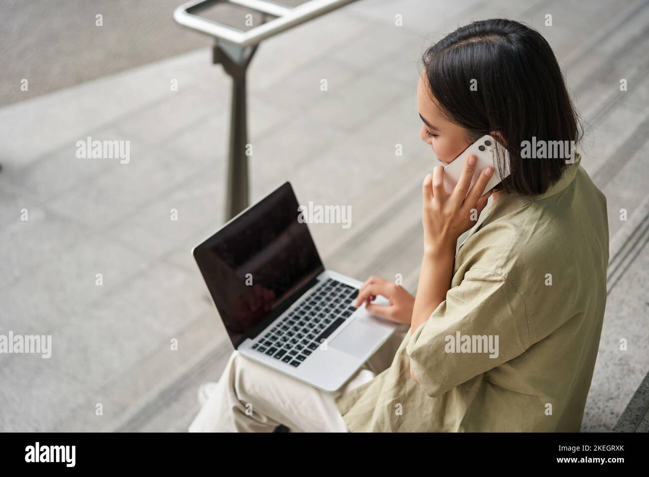 Portrait of asian girl, student sits on stairs with laptop, talks on ...
