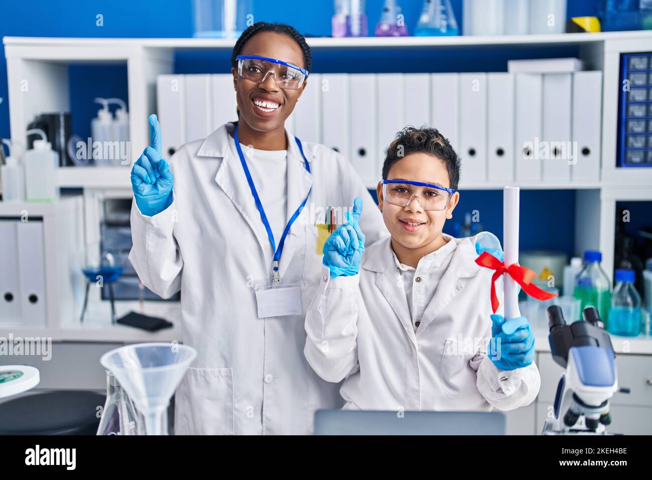 Mother and son at scientist laboratory smiling with an idea or question pointing finger with ...