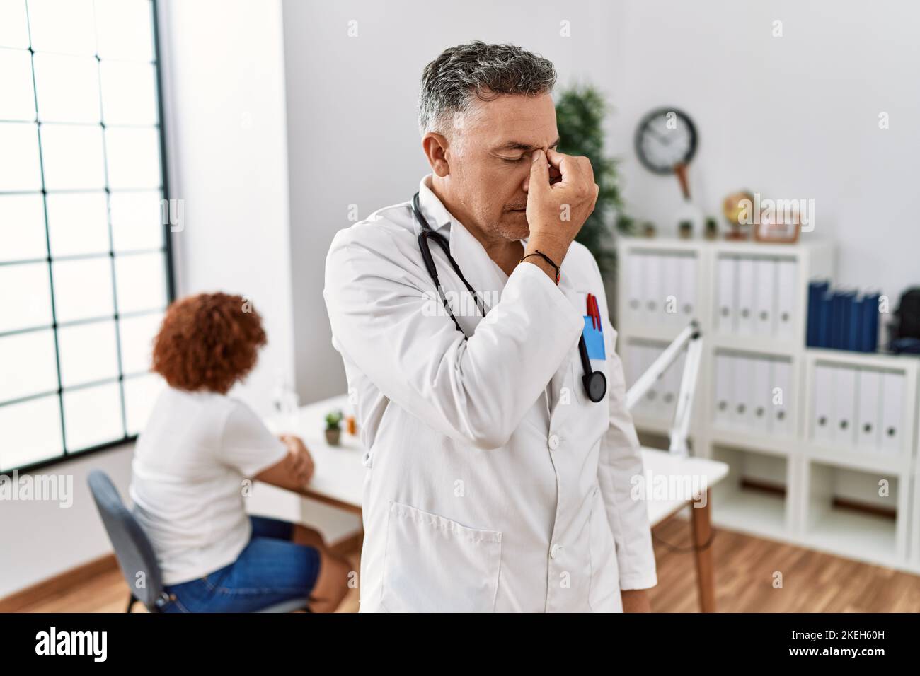 Middle age doctor man at the clinic with a patient tired rubbing nose ...