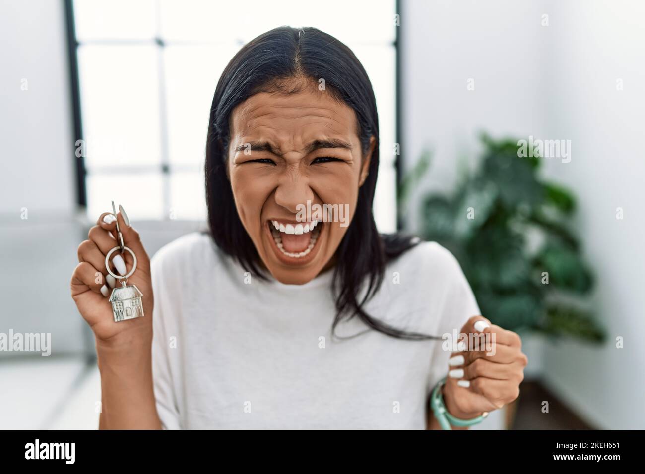 Young hispanic woman holding keys of new home angry and mad screaming frustrated and furious ...