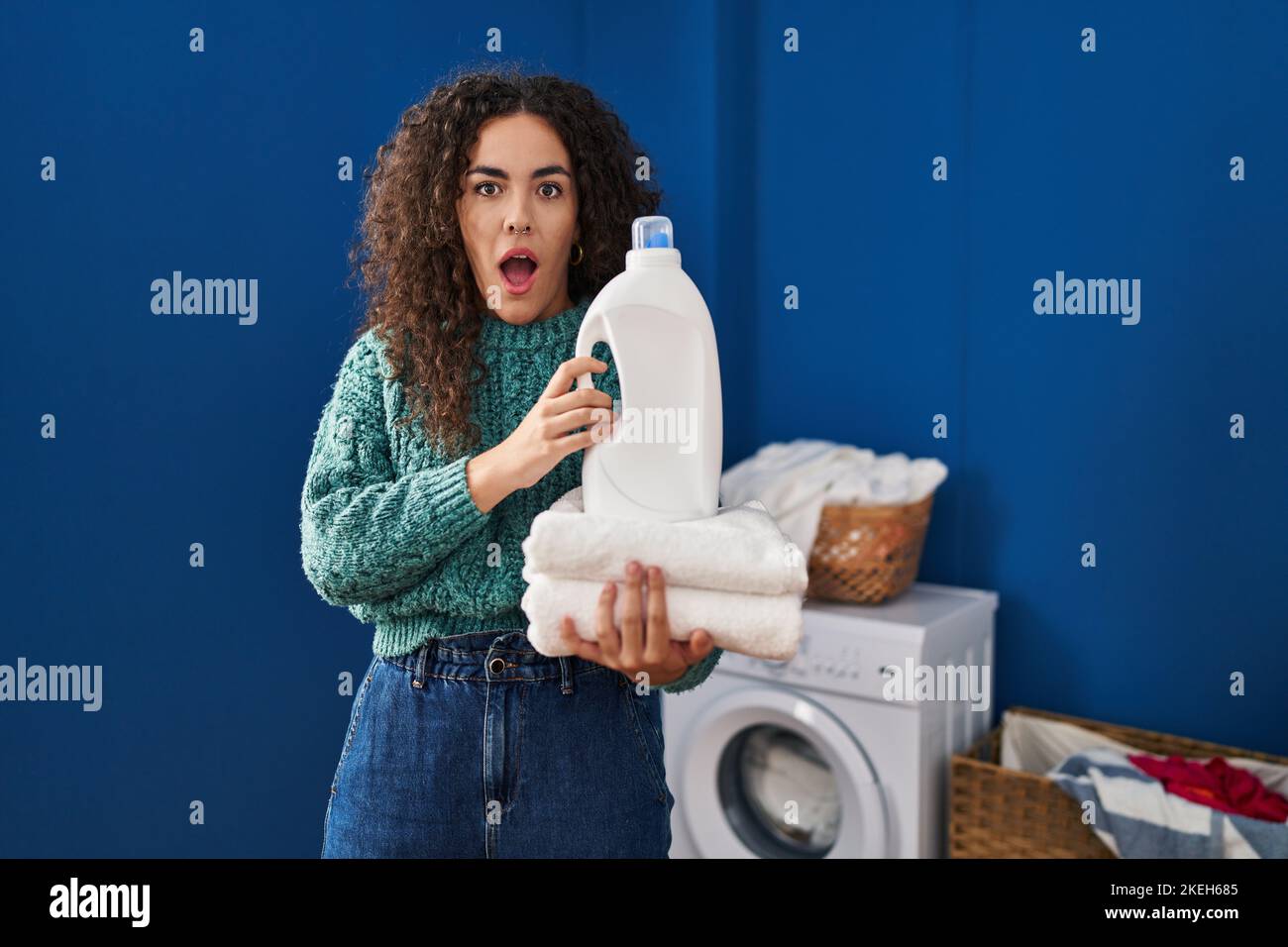 Young hispanic woman holding laundry and detergent bottle afraid and shocked with surprise and ...