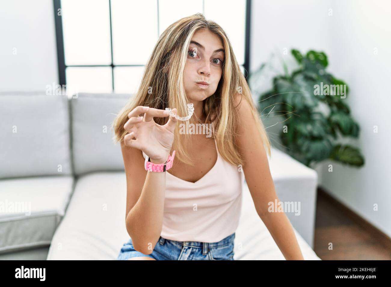 Young blonde woman holding invisible aligner orthodontic puffing cheeks ...