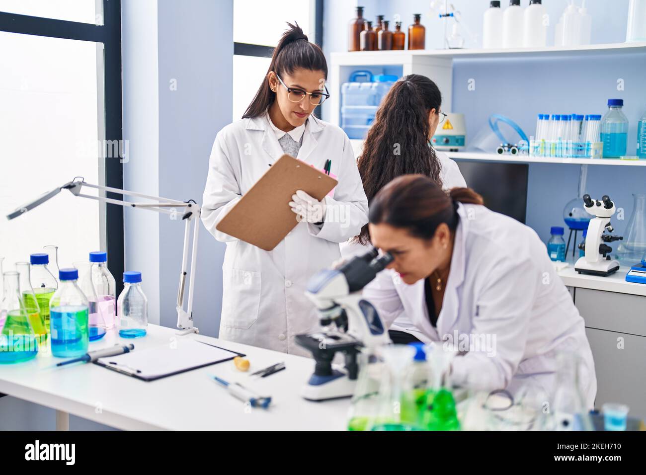 Three woman scientists using microscope write on checklist at ...