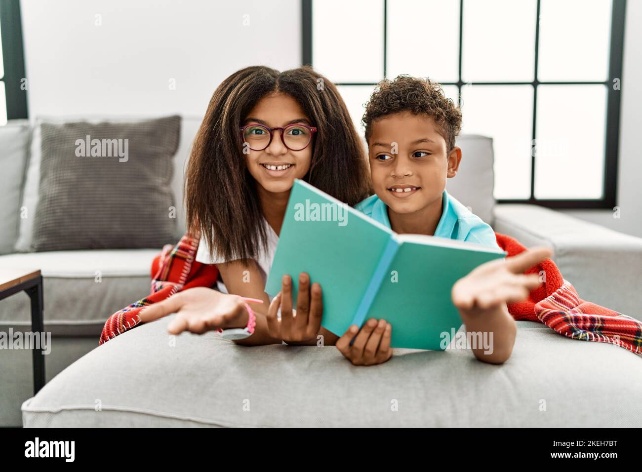 Two siblings lying on the sofa reading a book smiling cheerful offering ...