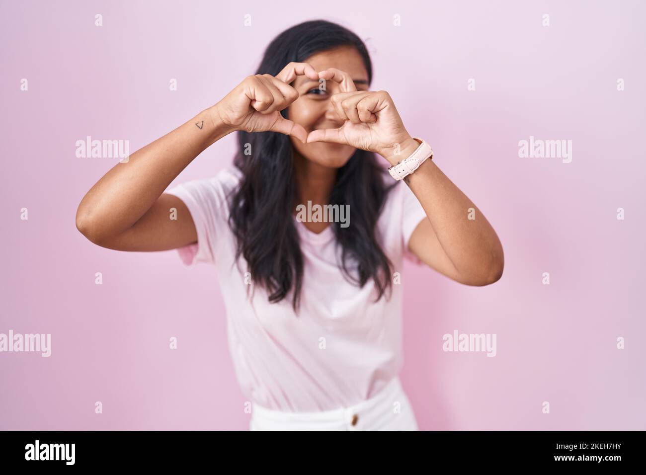 Young hispanic woman standing over pink background doing heart shape ...