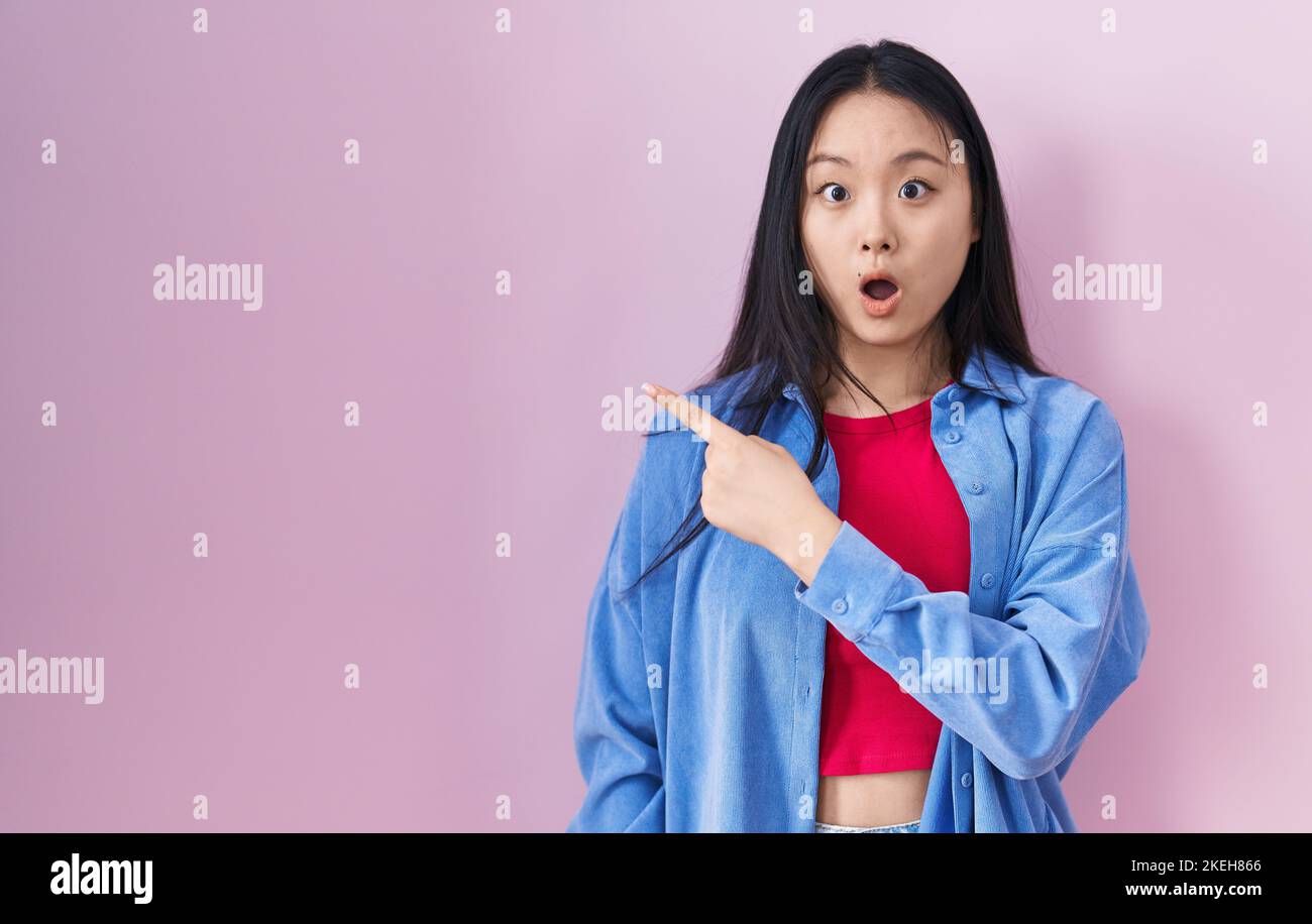 Young asian woman standing over pink background surprised pointing with ...