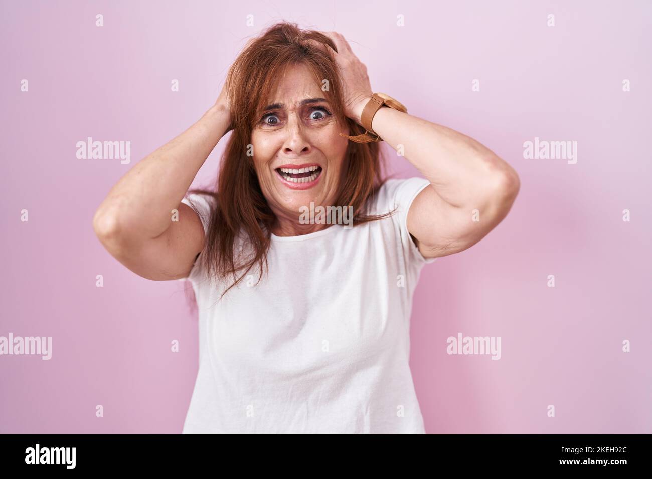 Middle age woman standing over pink background crazy and scared with ...