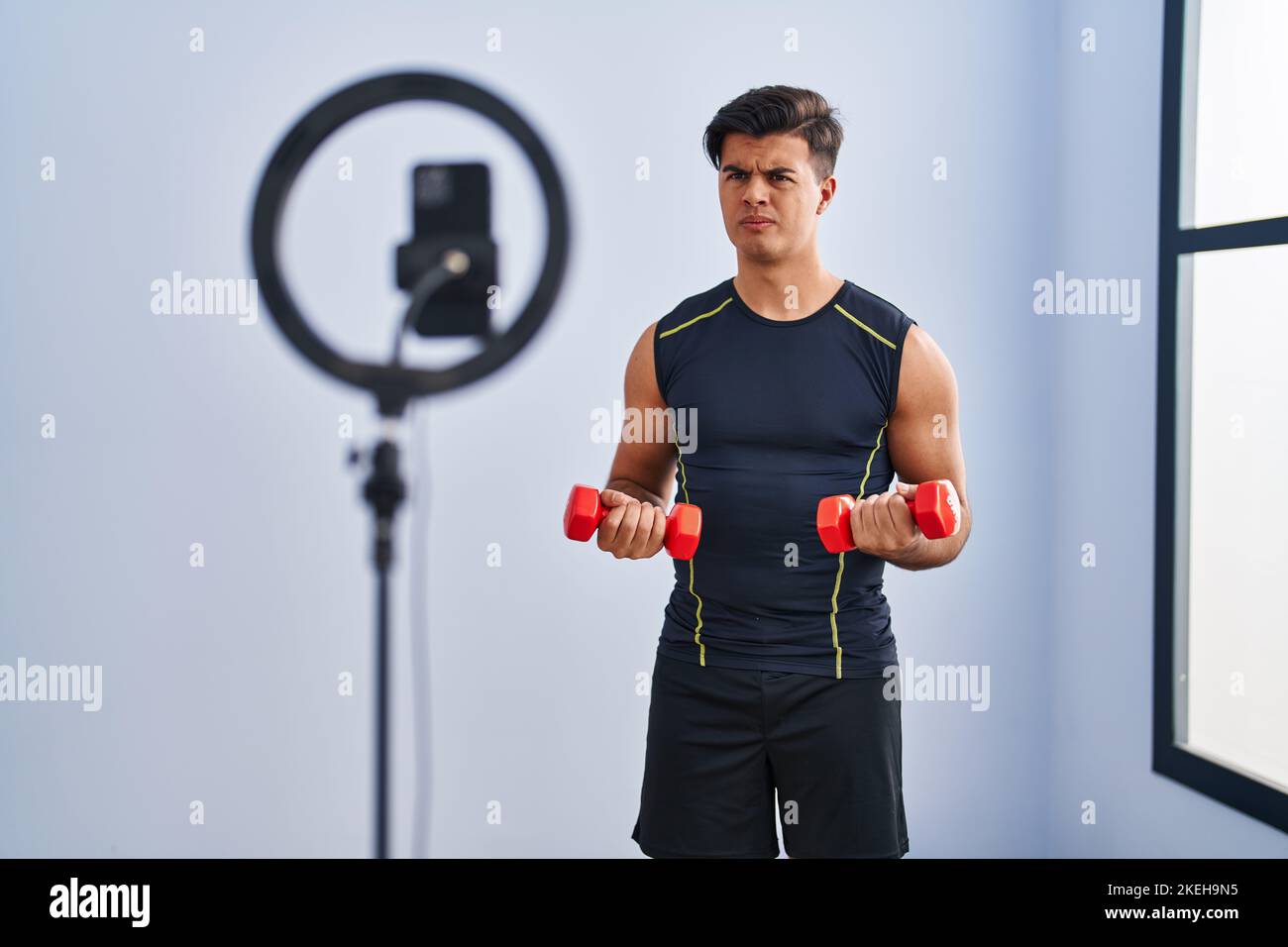 Hispanic man recording training tutorial at the gym skeptic and nervous, frowning upset because ...