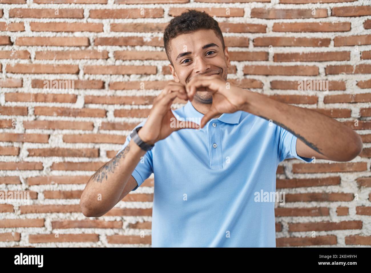 Brazilian young man standing over brick wall smiling in love doing ...
