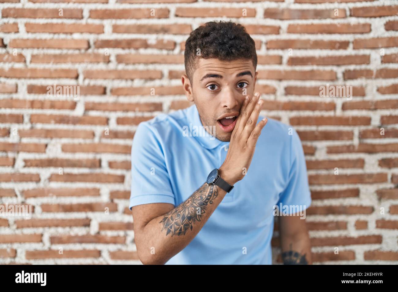 Brazilian young man standing over brick wall hand on mouth telling ...