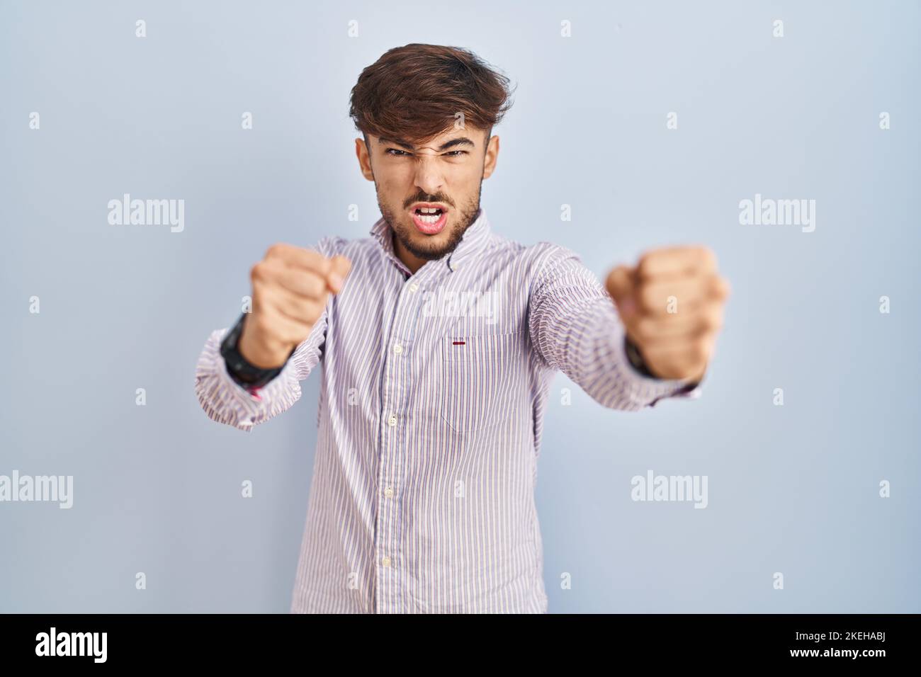Arab man with beard standing over blue background angry and mad raising ...