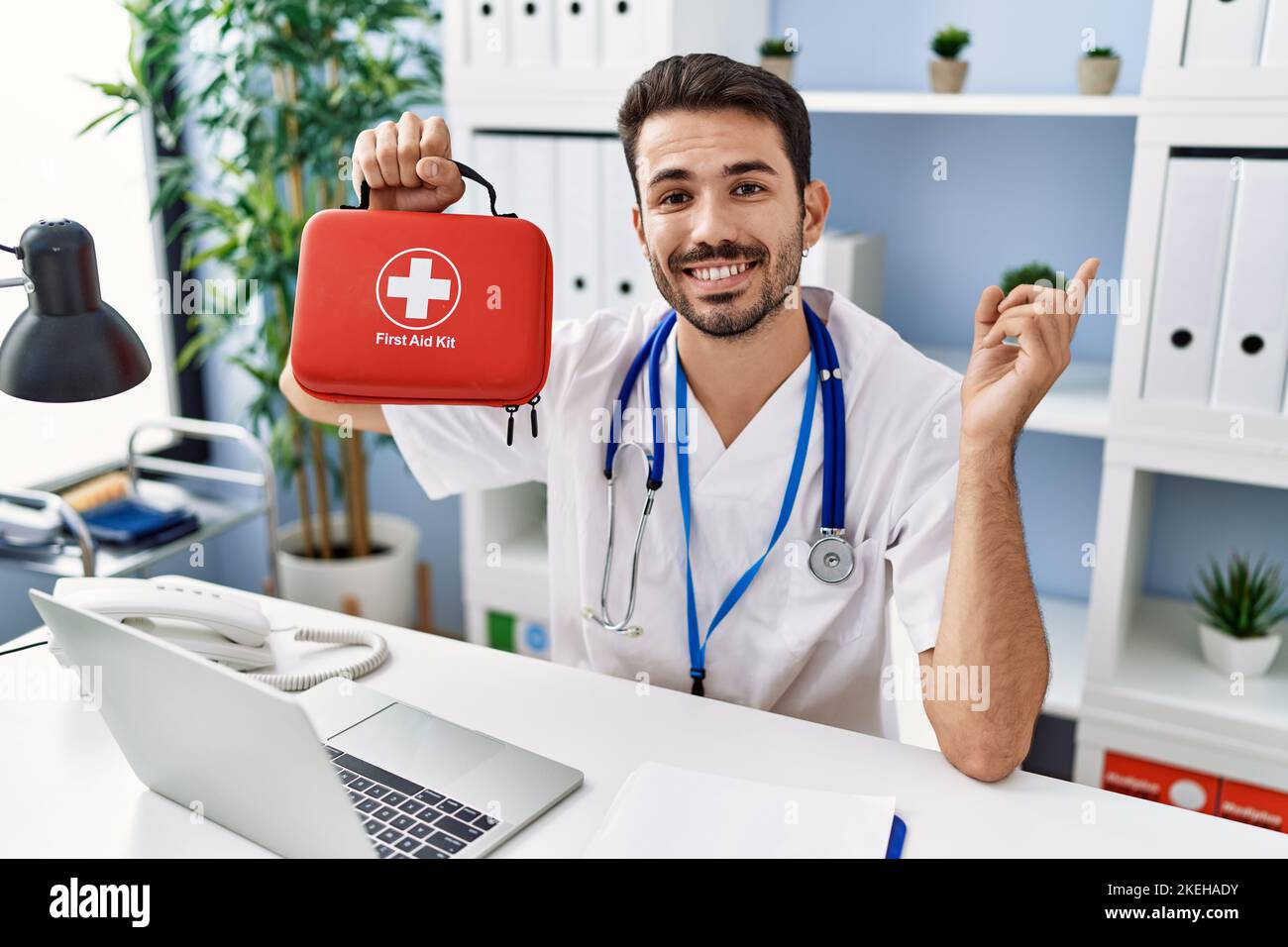 Young hispanic doctor man holding first aid kit smiling happy pointing ...