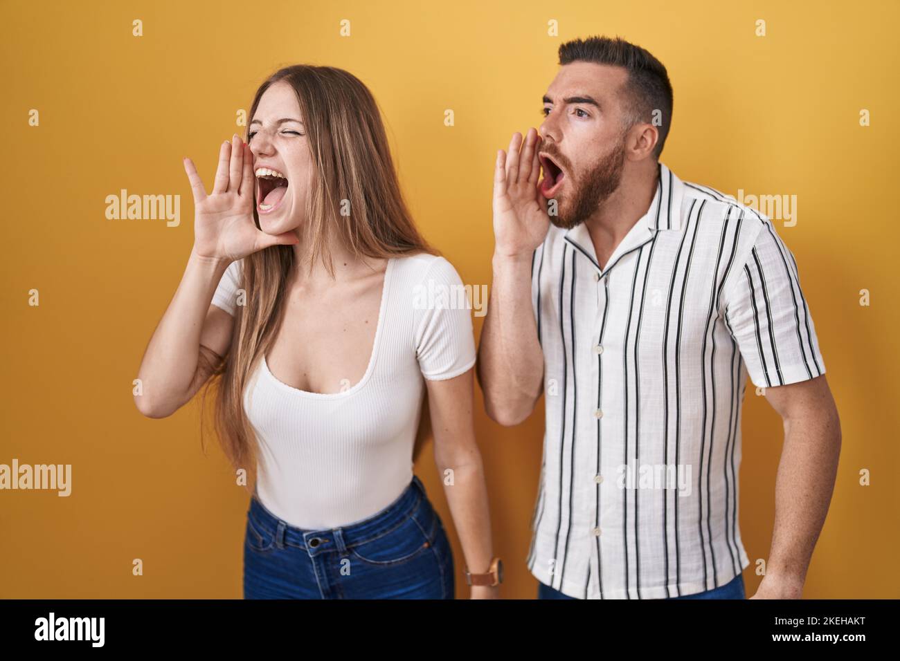 Young couple standing over yellow background shouting and screaming ...