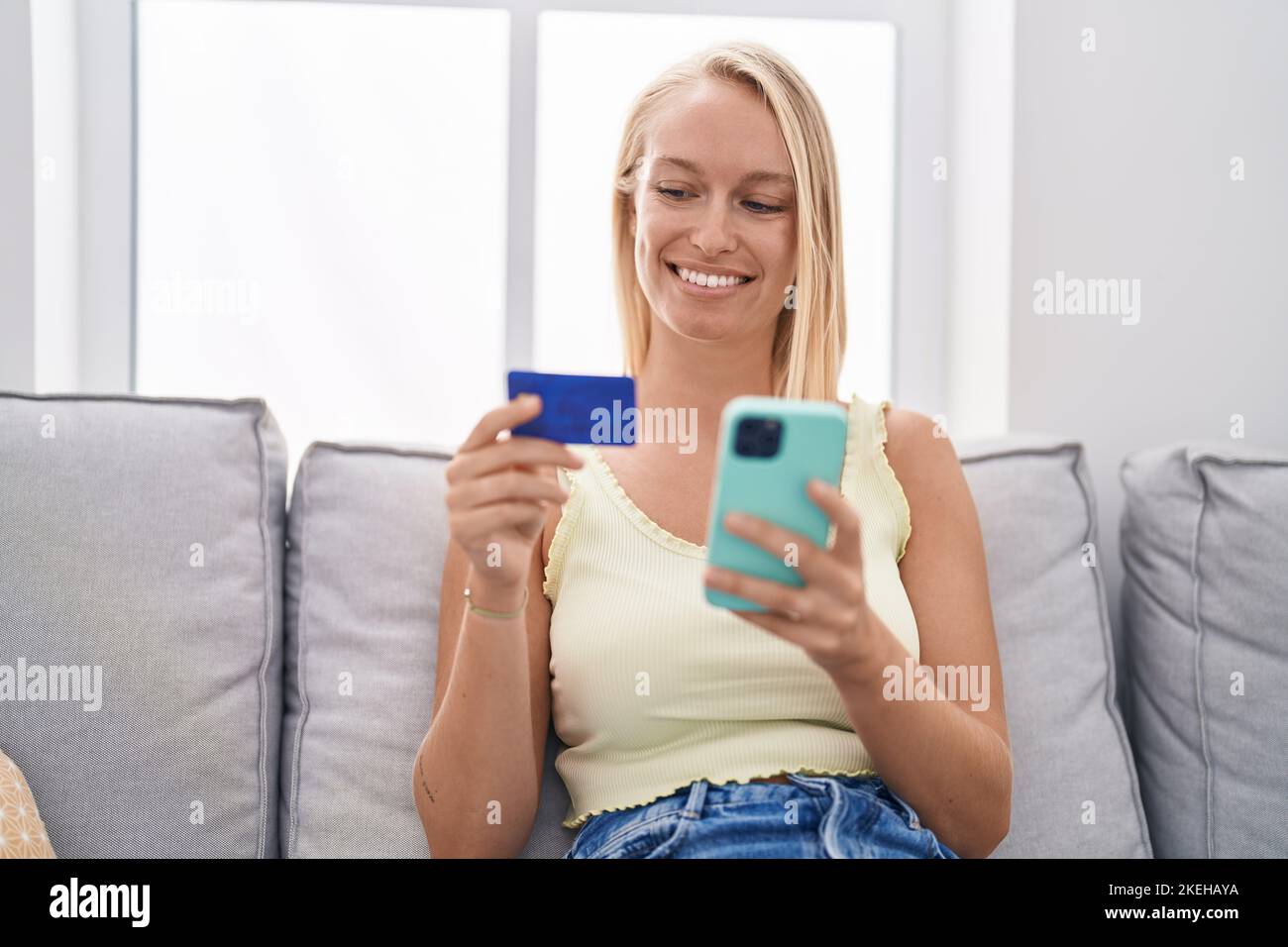 Young blonde woman using smartphone and credit card sitting on sofa at ...