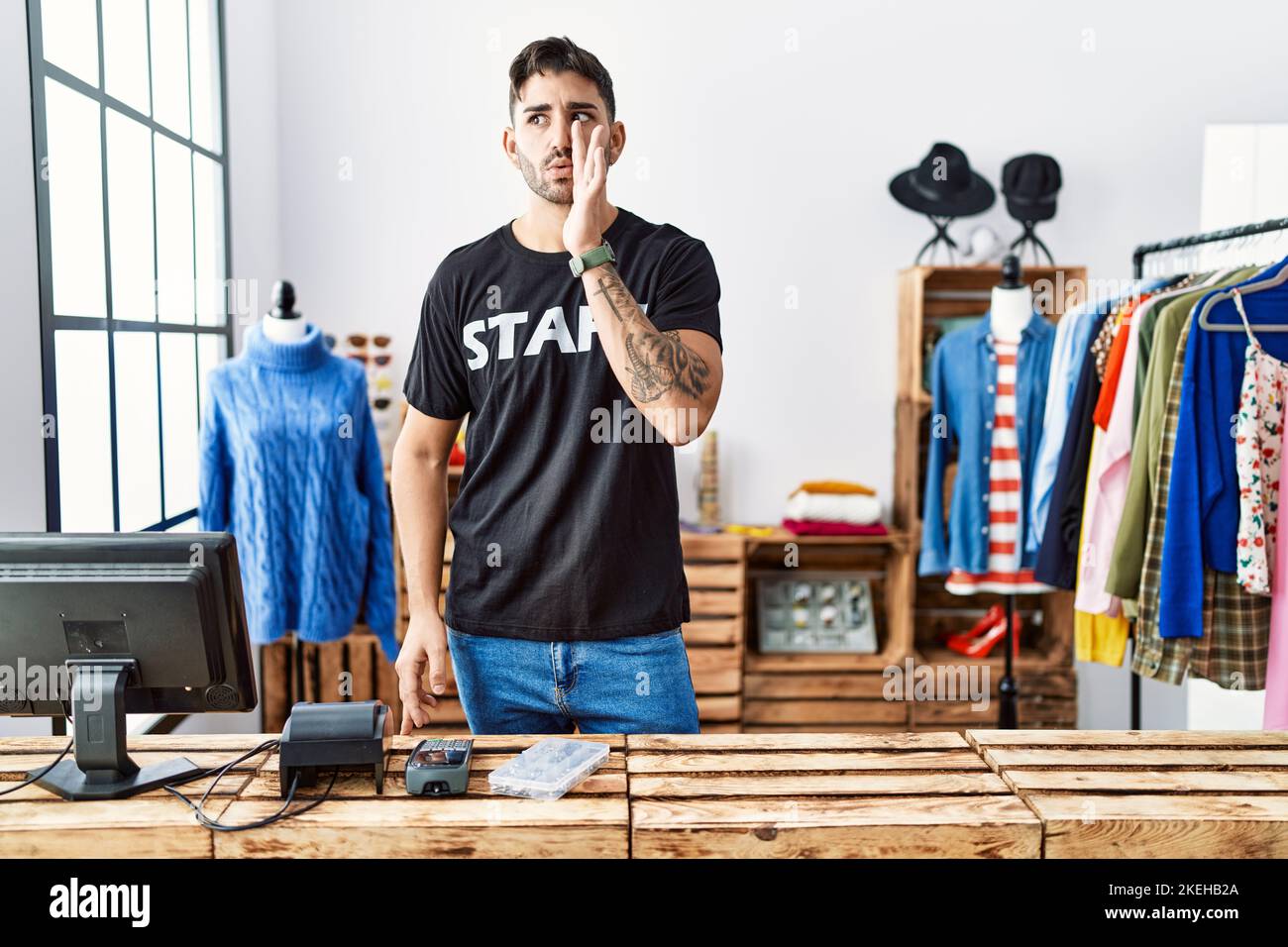 Young hispanic man working at retail boutique hand on mouth telling ...