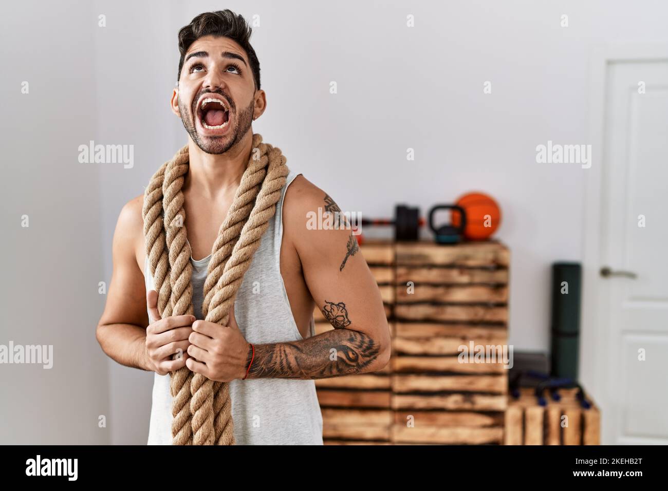 Young hispanic man training with battle rope at the gym angry and mad ...