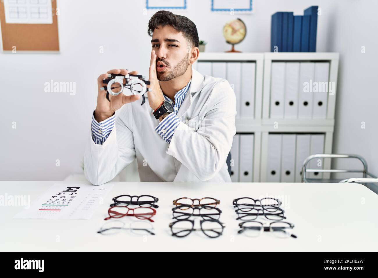 Young optician man holding optometry glasses hand on mouth telling ...