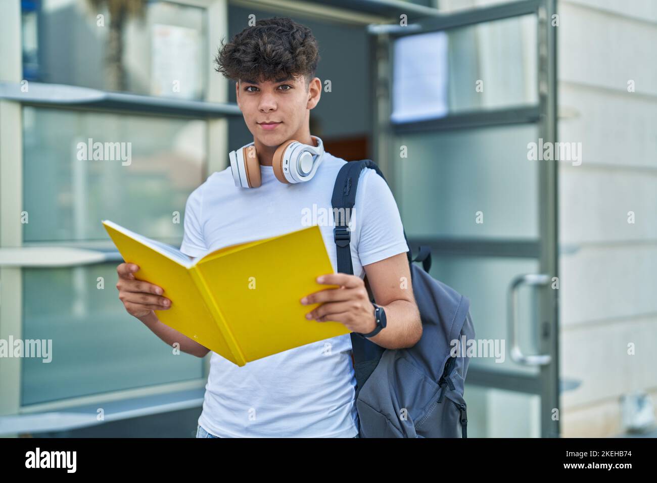 Young hispanic teenager student wearing backpack reading book at ...