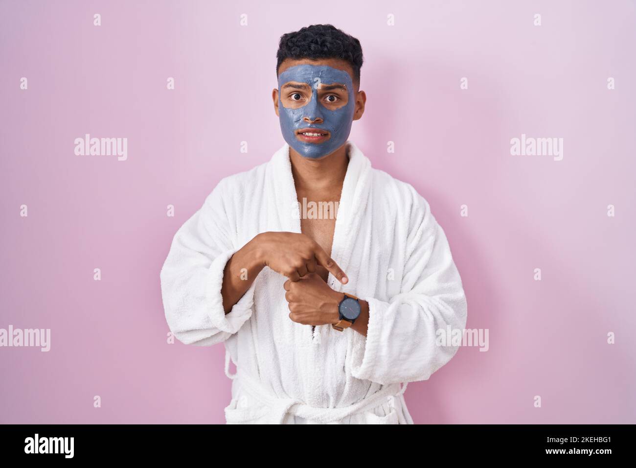 Young hispanic man wearing beauty face mask and bath robe in hurry ...