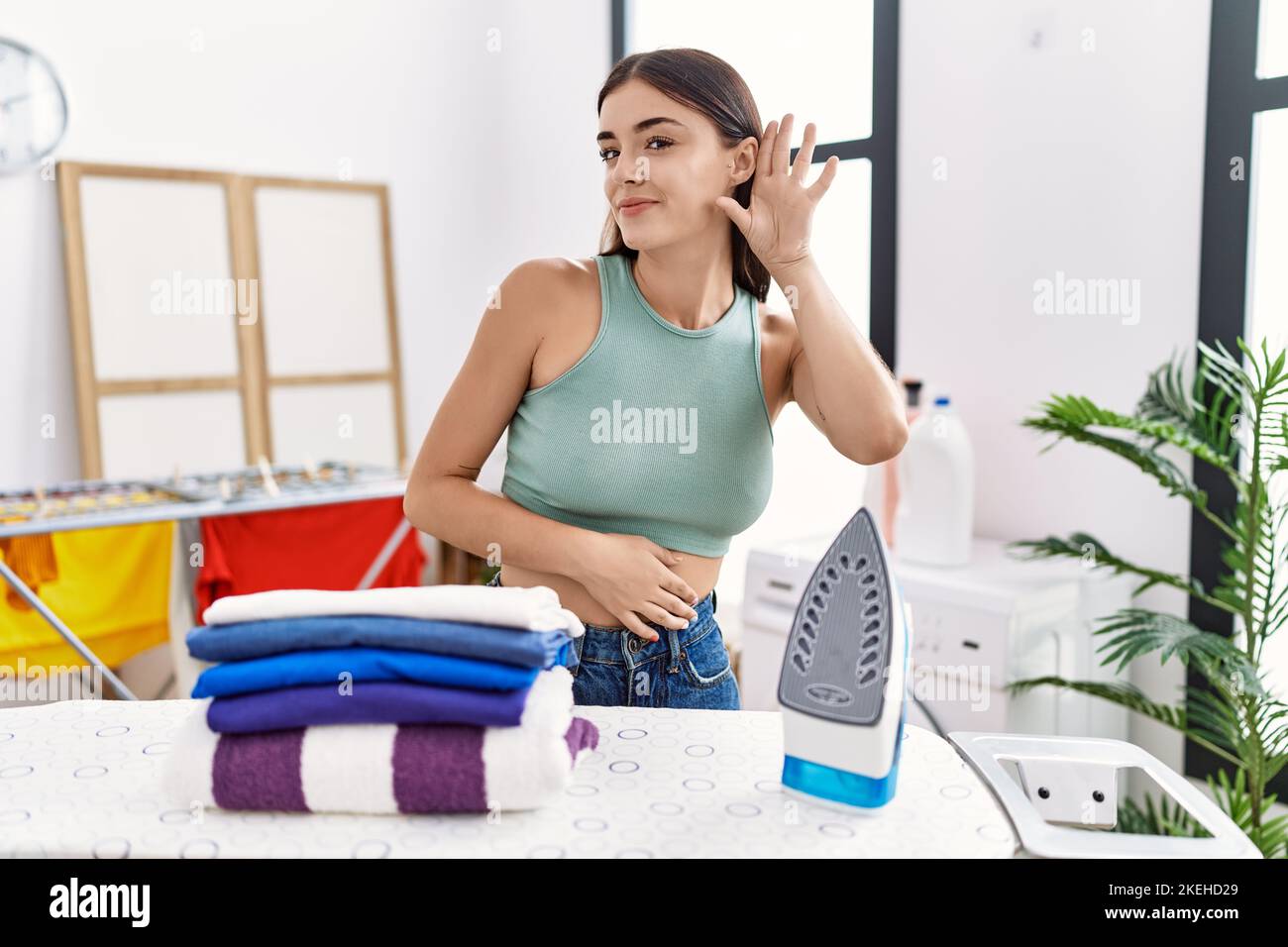 Young hispanic woman ironing clothes at laundry room smiling with hand ...