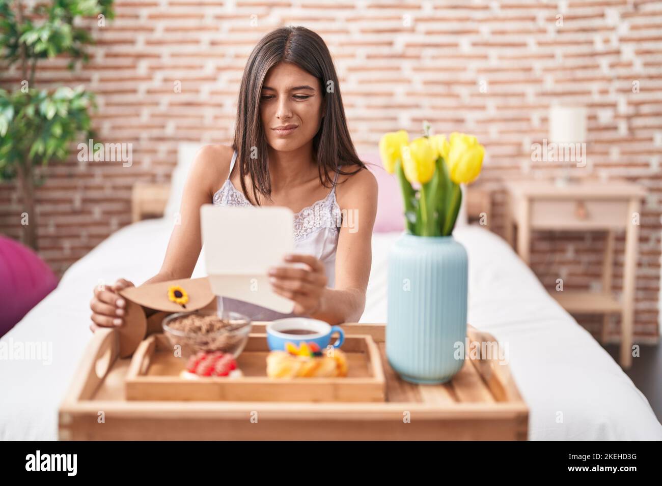Brunette young woman eating breakfast in the bed reading a letter ...