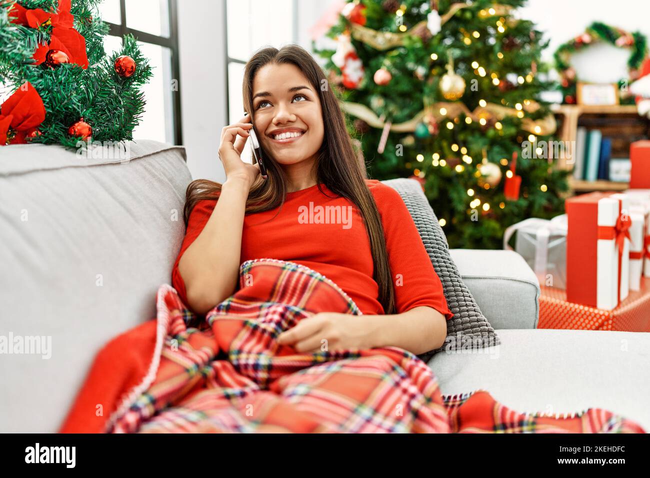 Young latin woman talking on the smartphone sitting by christmas tree ...