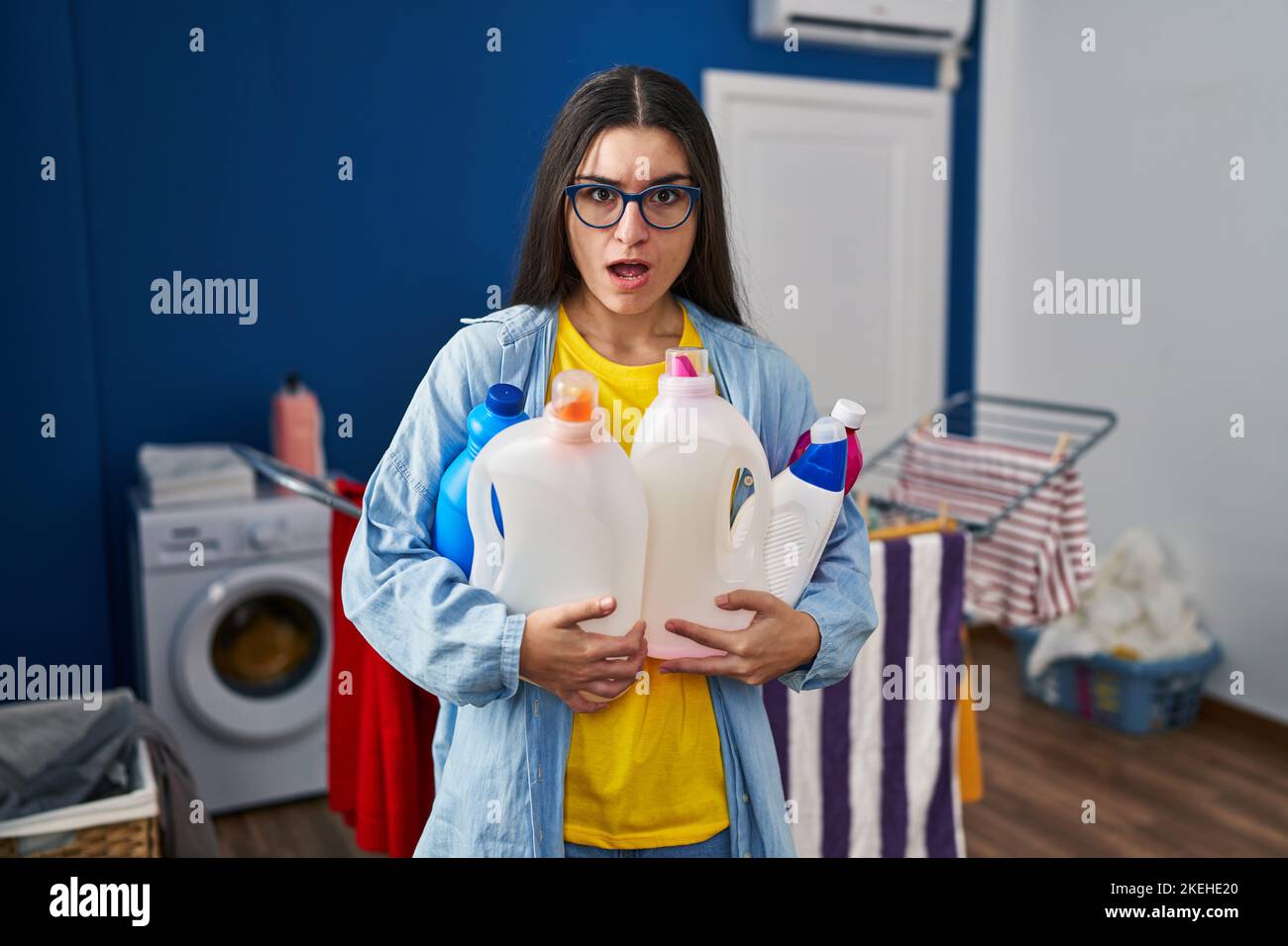 Young hispanic woman holding detergent bottles in shock face, looking ...