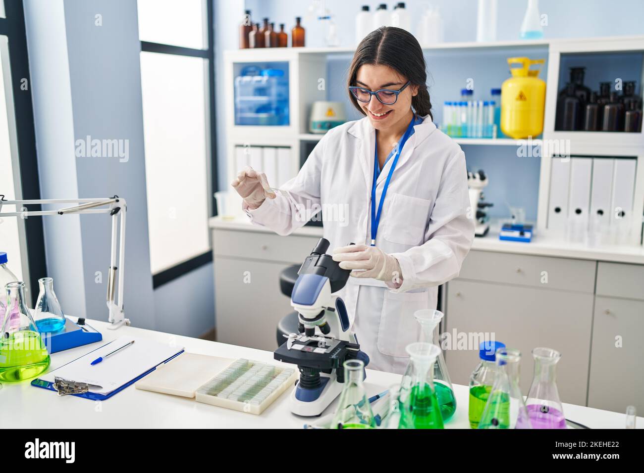 Young hispanic woman wearing scientist uniform using microscope at ...