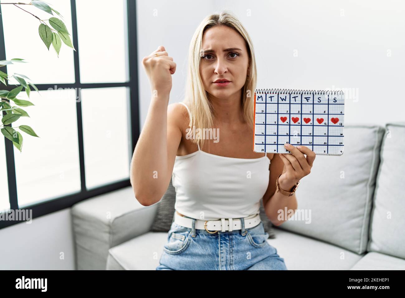 Young blonde woman holding heart calendar angry and mad raising fist ...