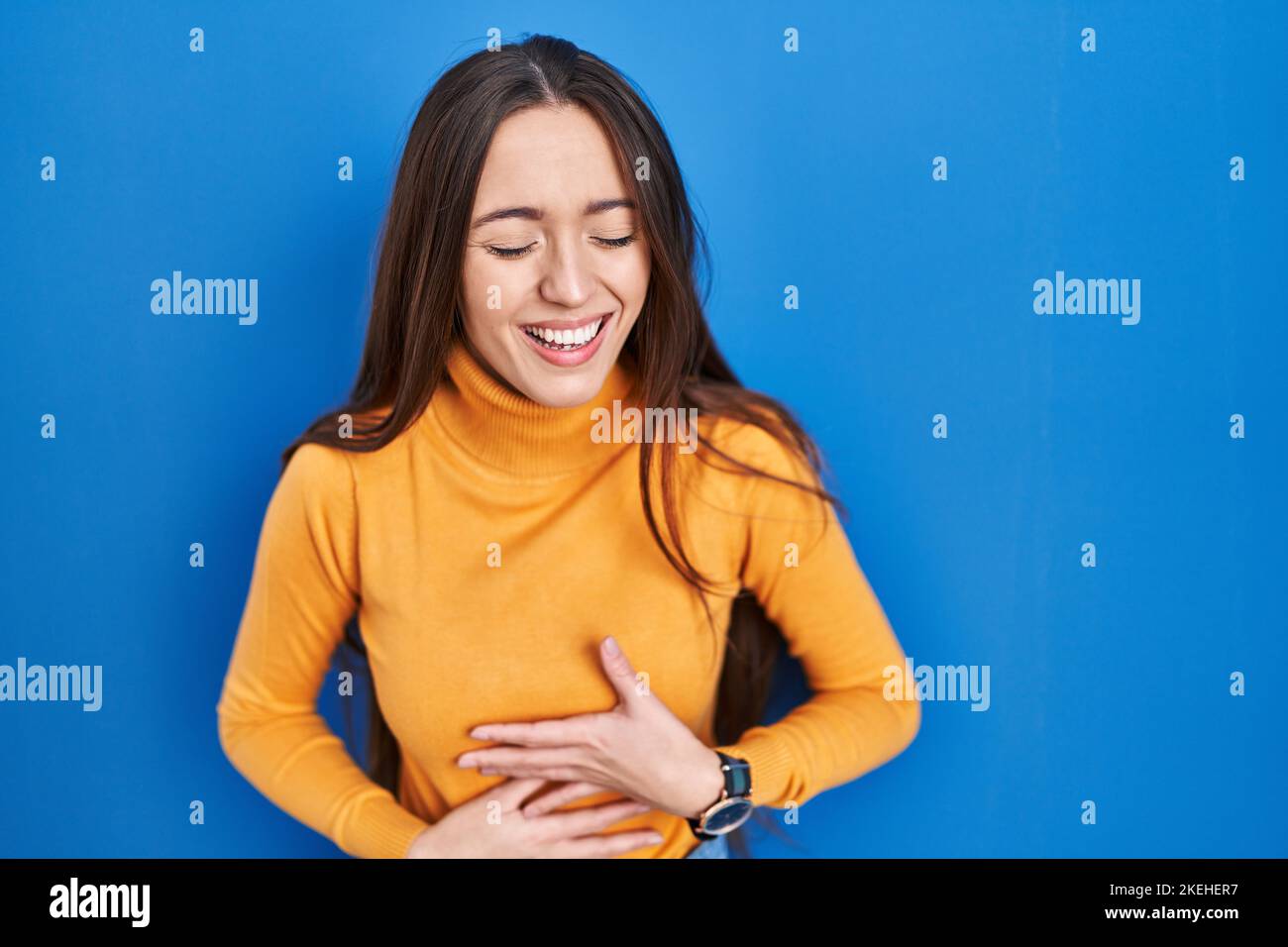 Young brunette woman standing over blue background smiling and laughing hard out loud because ...