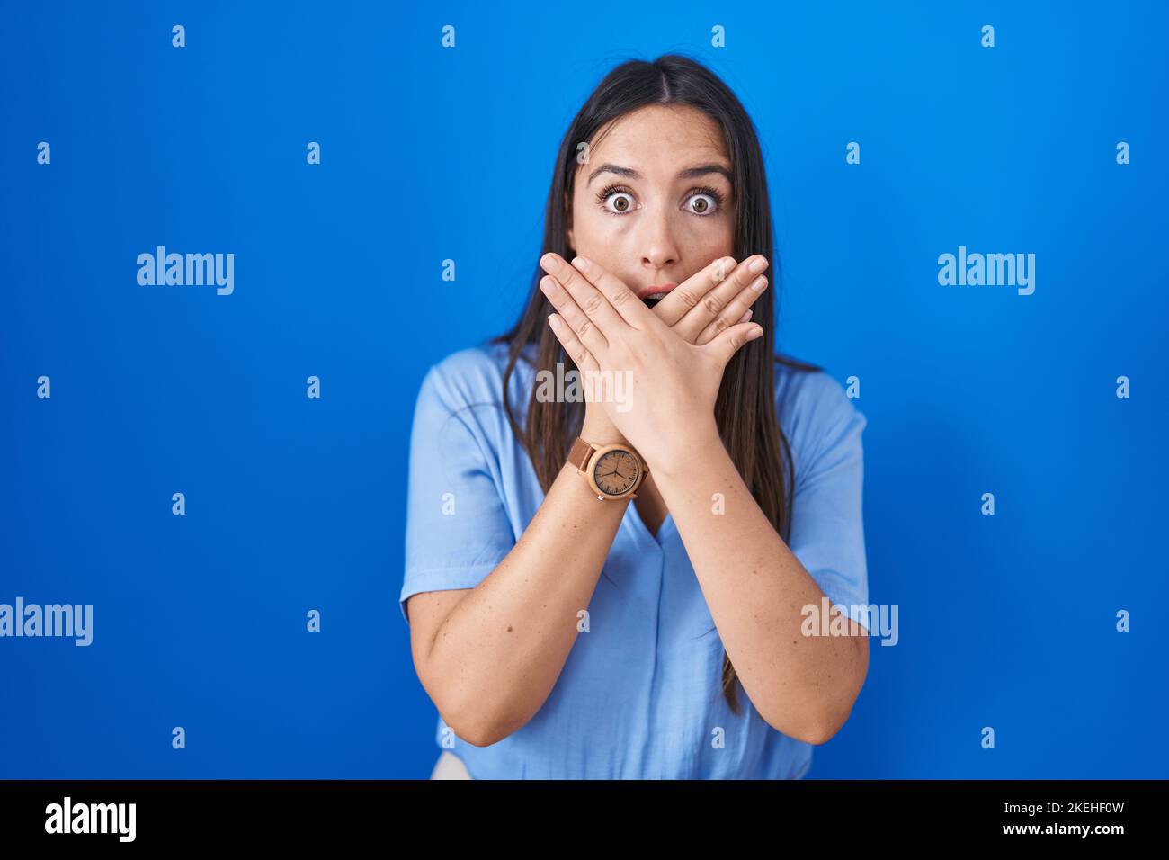 Young brunette woman standing over blue background shocked covering ...