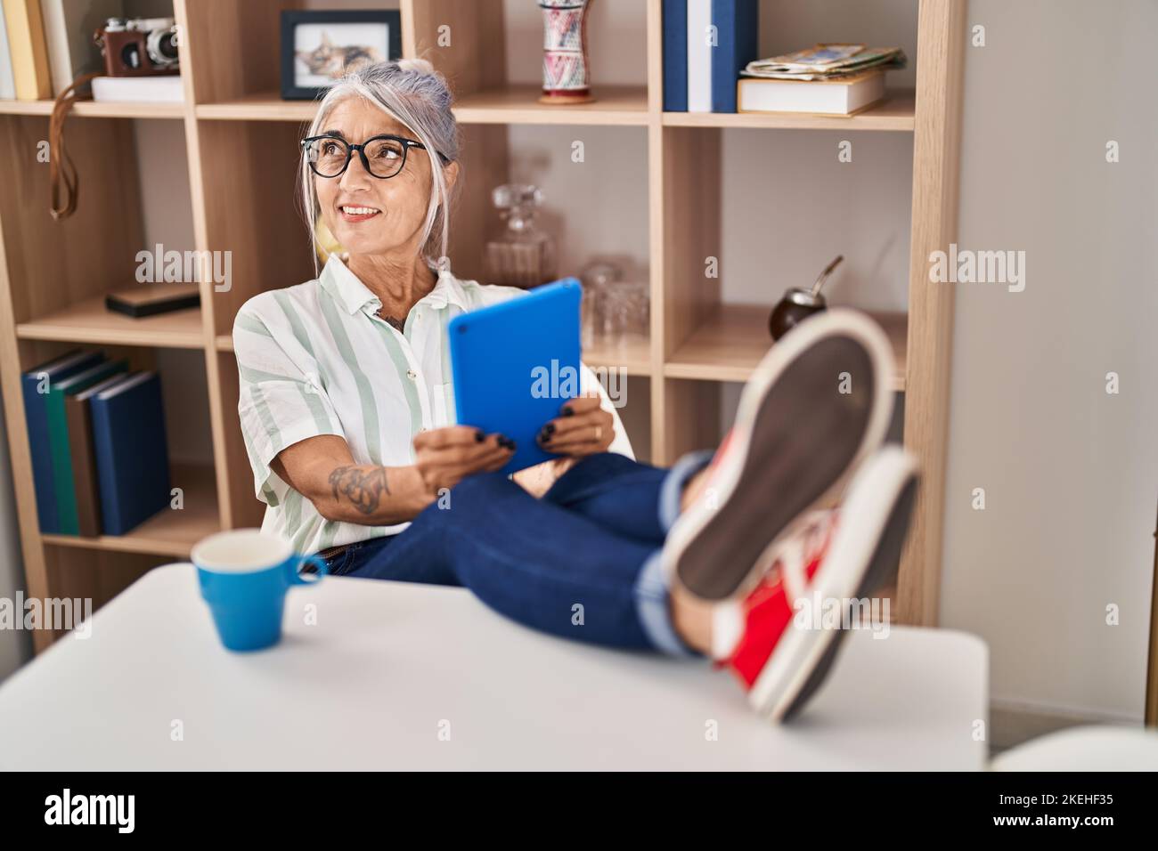 Middle age grey-haired woman drinking coffee and using touchpad at home ...