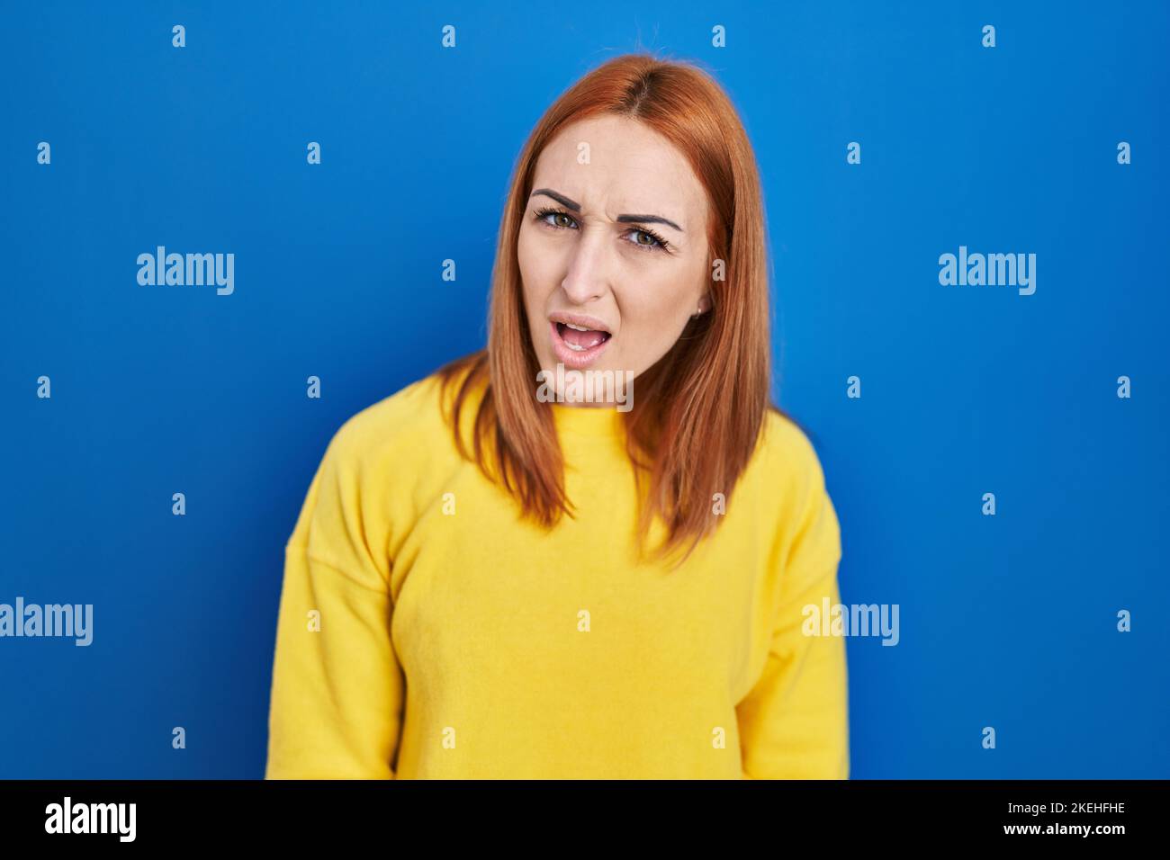 Young woman standing over blue background in shock face, looking ...