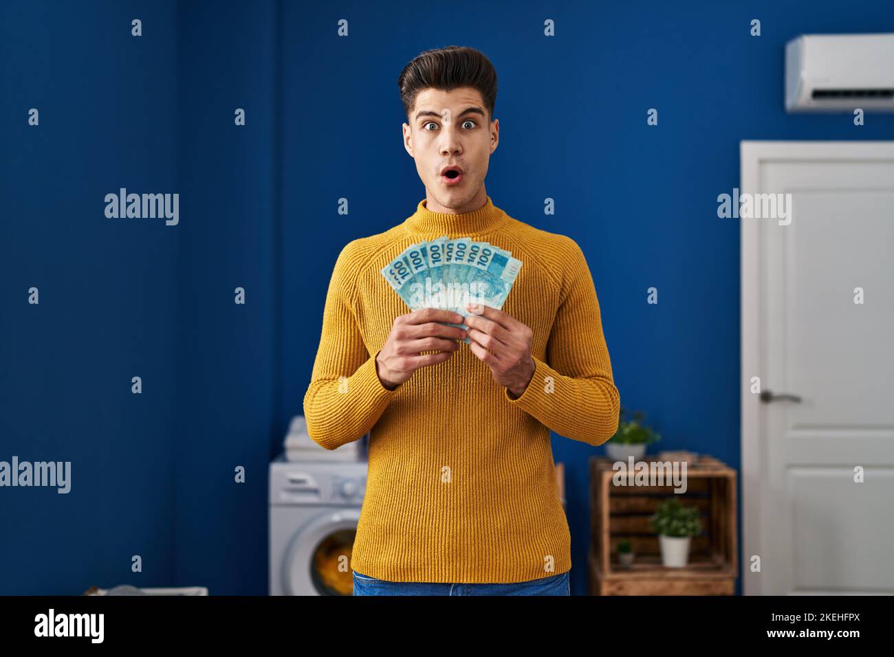 Young hispanic man at laundry room holding brazilian reals afraid and ...