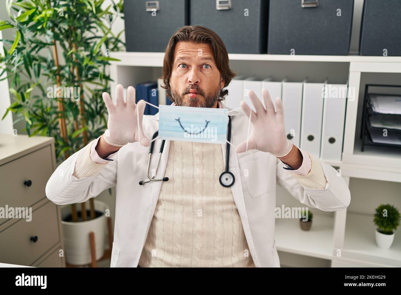 Handsome middle age doctor man wearing safety mask making fish face ...