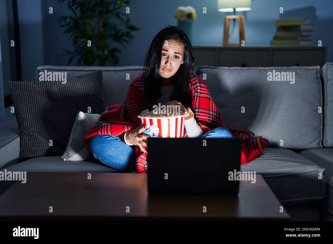 Hispanic woman eating popcorn watching a movie on the sofa depressed ...