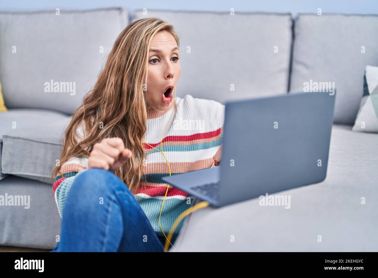 Young blonde woman using laptop at home scared and amazed with open ...