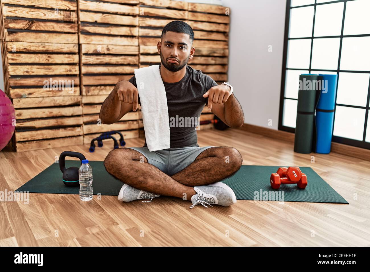 Young indian man sitting on training mat at the gym pointing down ...