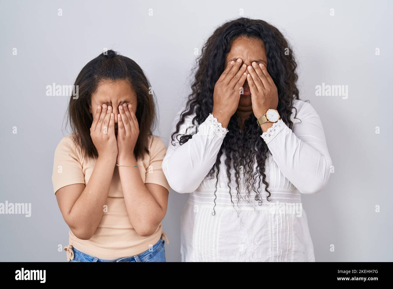 Mother and young daughter standing over white background rubbing eyes ...