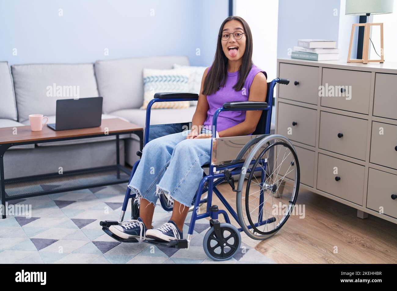 Young hispanic woman sitting on wheelchair at home sticking tongue out ...